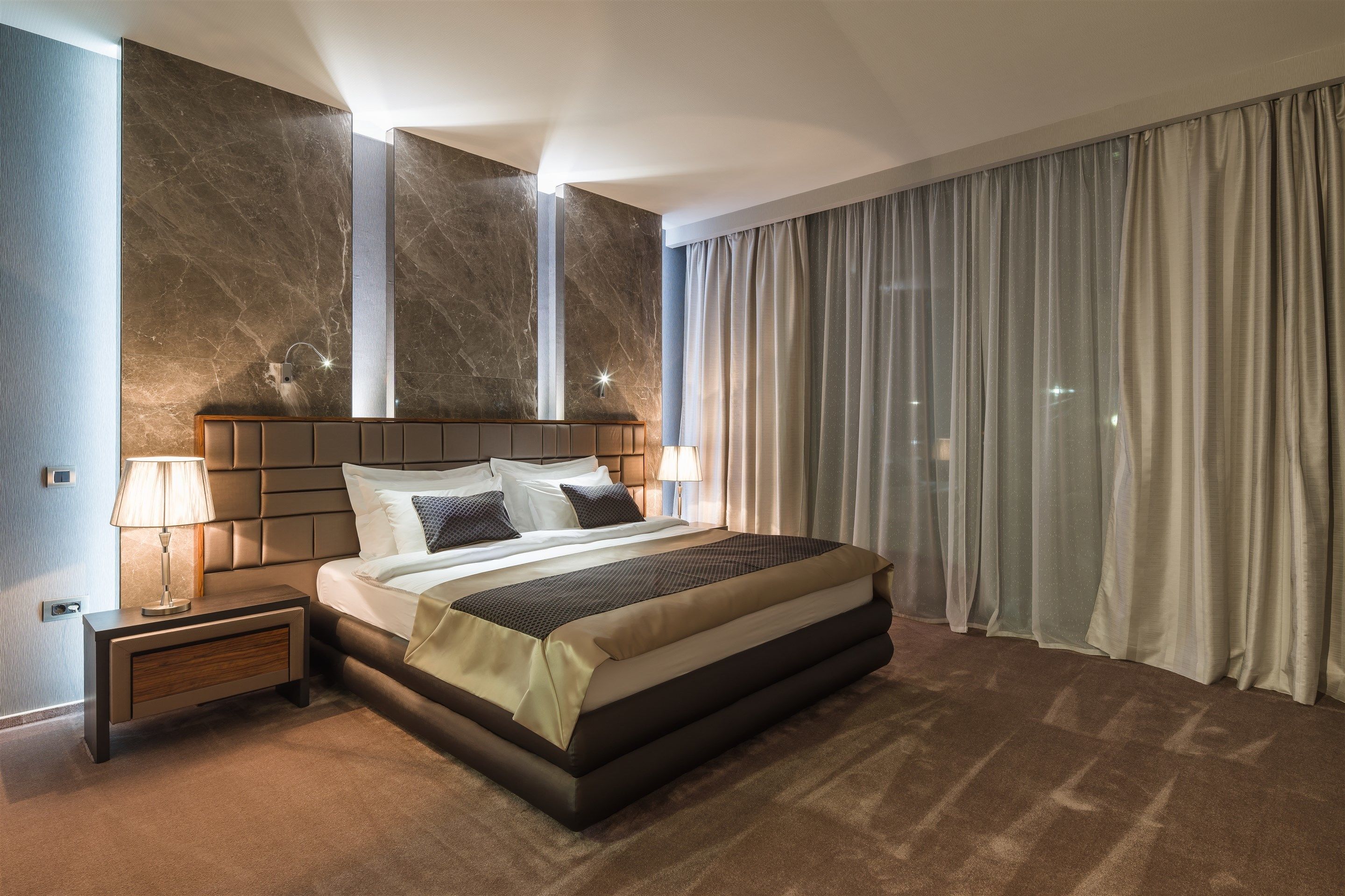Quarto de hotel moderno com uma cama grande, uma mesa de cabeceira com abajur, parede de mármore e vista da cidade através de cortinas leves.