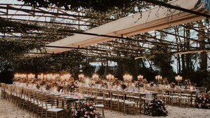 Espaço para eventos ao ar livre com mesas preparadas para uma recepção, rodeado por uma estrutura de pérgula e decorado com arranjos florais.