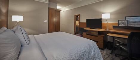 Quarto de hotel com uma cama grande, uma escrivaninha com computador, uma TV e uma cadeira.