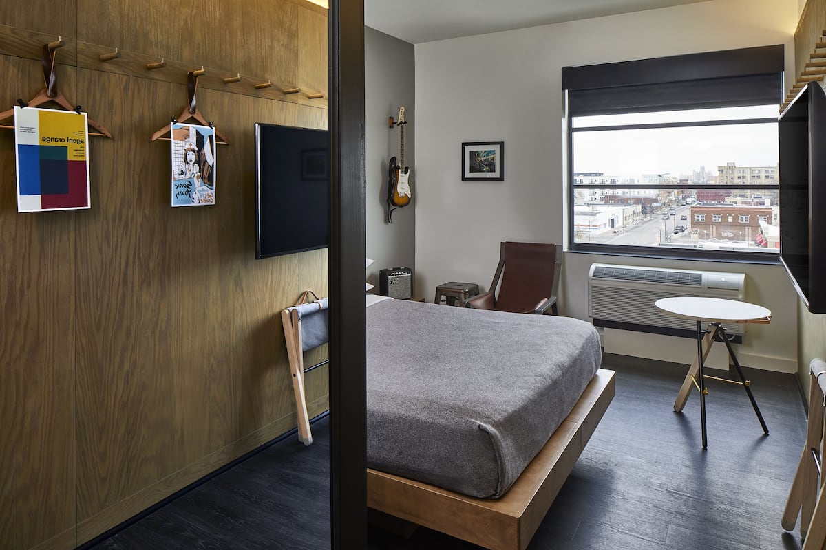 Quarto de hotel moderno com uma cama, uma pequena mesa, uma cadeira, um violão e uma janela com vista para a cidade.