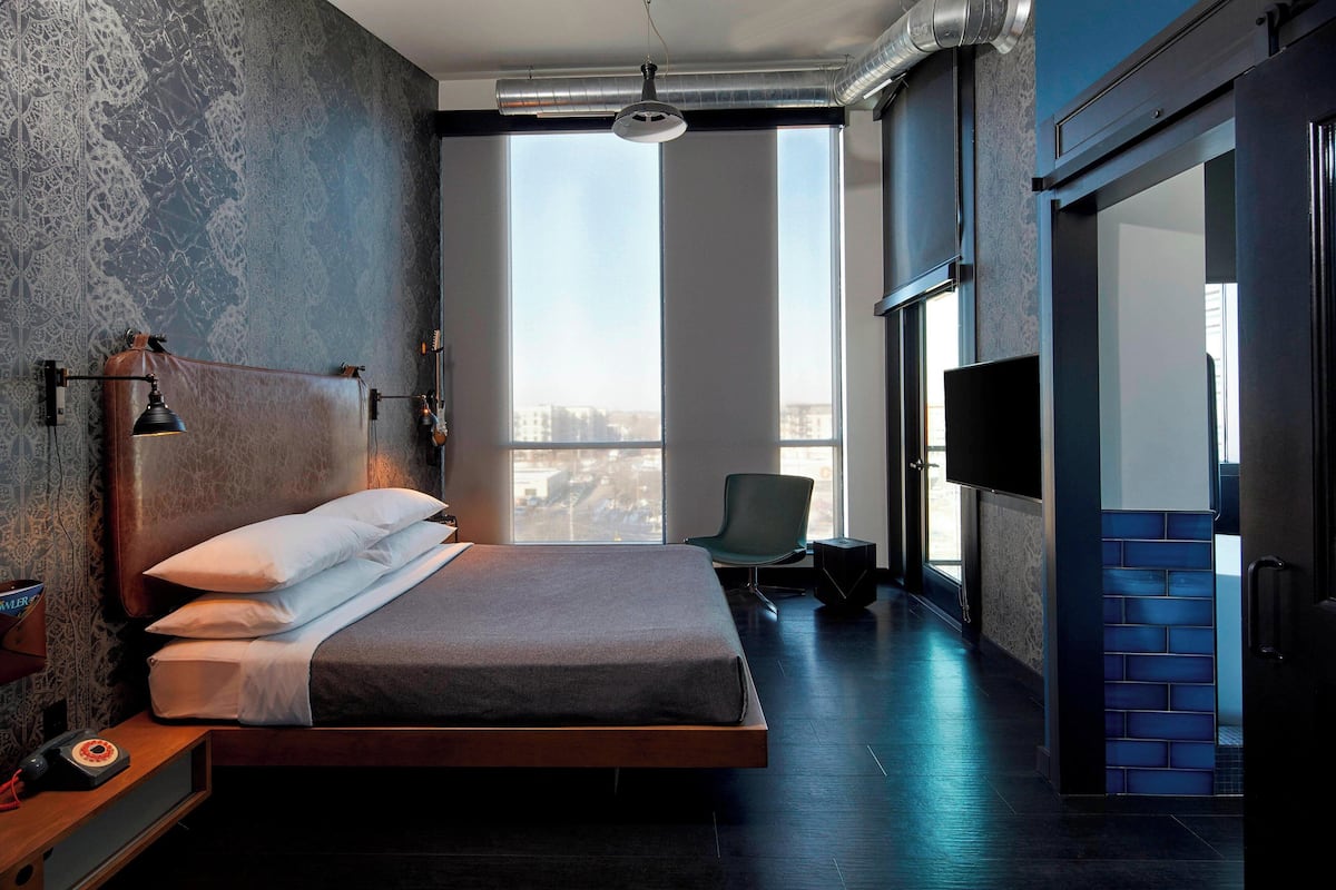 Quarto de hotel moderno com uma cama grande, vista para a cidade, televisão e uma cadeira.