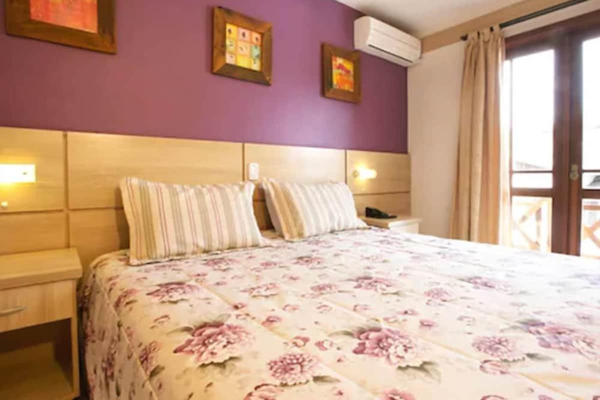 Quarto com cama, cabeceira, mesas de cabeceira e luzes fixadas na parede.
