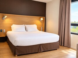 Quarto de hotel com uma cama grande, cabeceira de madeira, duas luminárias fixadas na parede, janela com cortinas e piso de madeira.