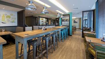 Restaurante moderno com mesas de madeira compridas, banquetas e iluminação pendente.