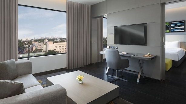 Quarto de hotel moderno com uma grande janela, televisão de tela plana, escrivaninha, cadeira, sofá e cama.