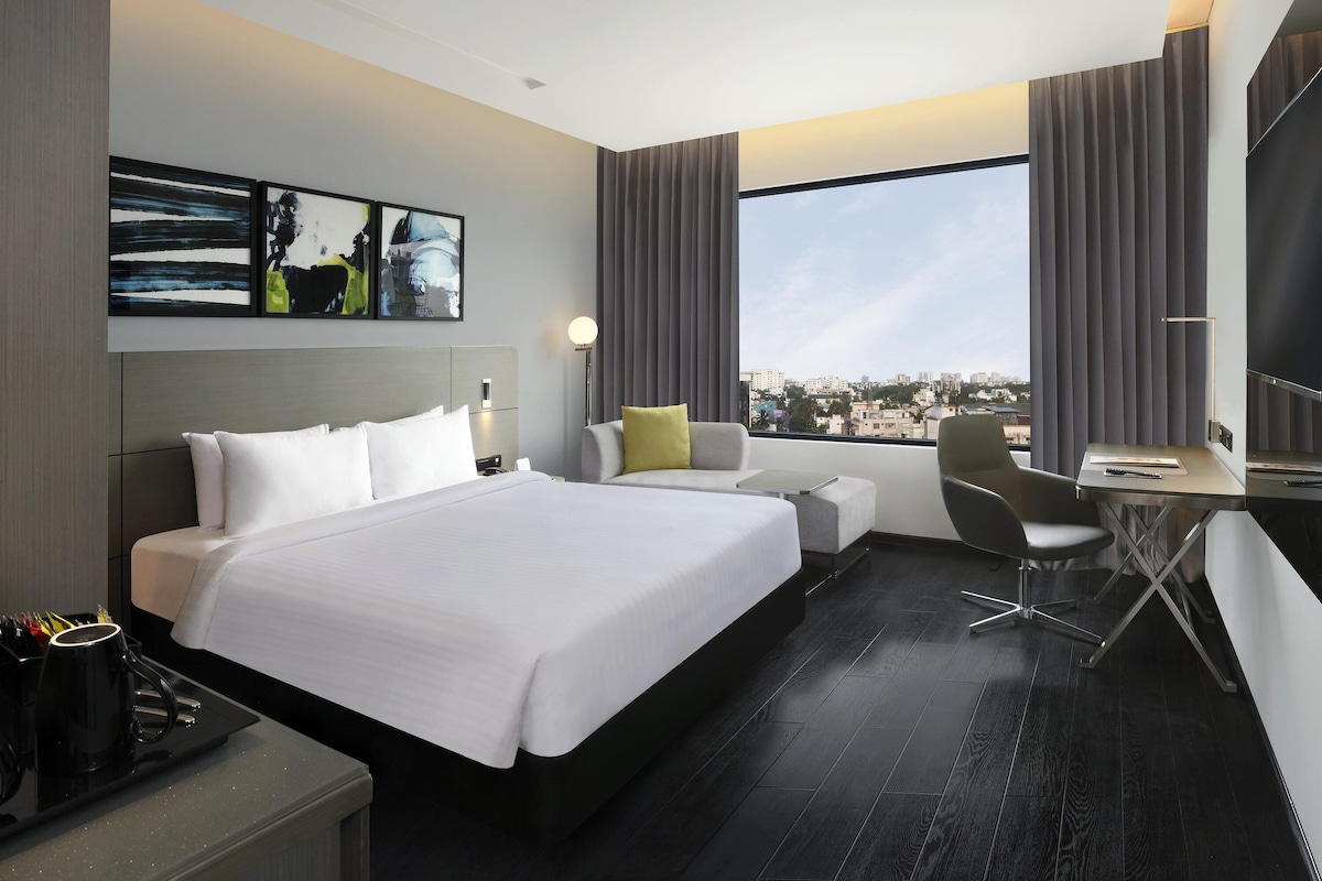 Quarto de hotel moderno com uma cama grande, uma escrivaninha e uma cadeira. Vê-se uma vista da cidade pela janela, e o quarto tem um design minimalista.