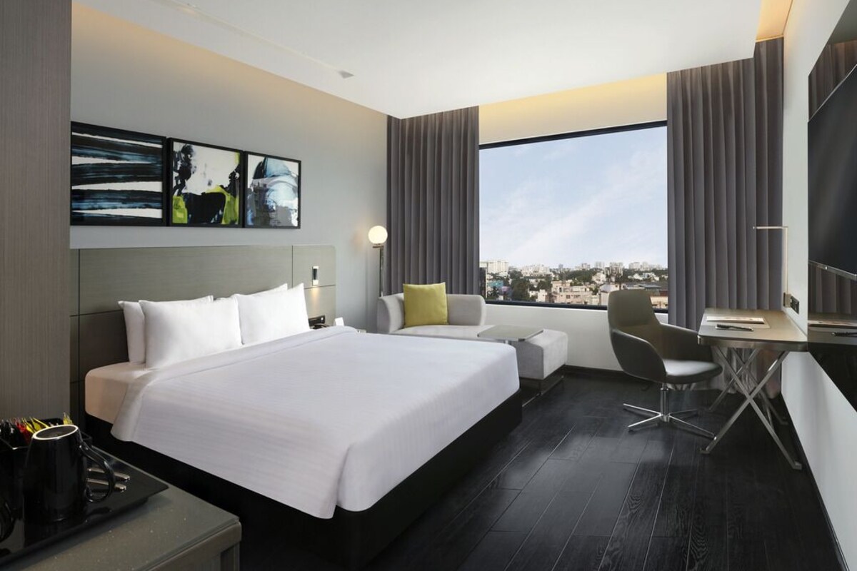 Quarto de hotel moderno com uma cama grande, uma escrivaninha, uma cadeira e vista para a cidade.