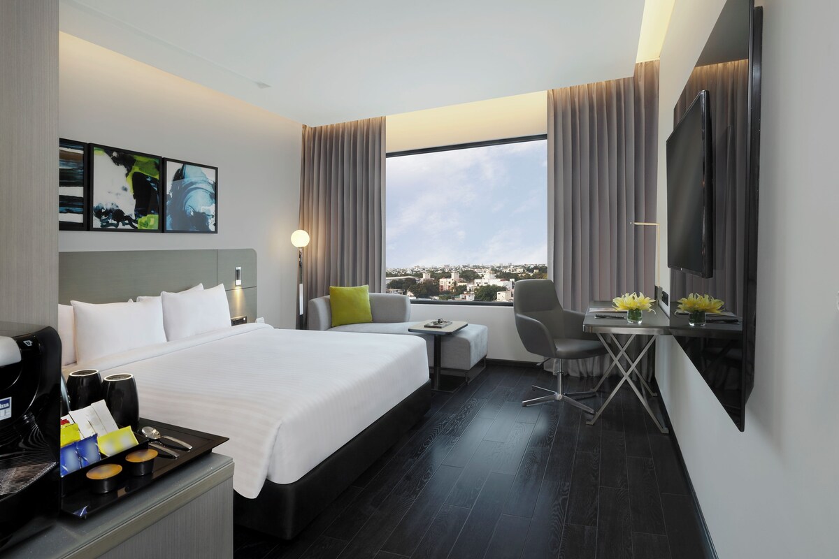 Quarto de hotel com cama, escrivaninha, cadeira, televisão e janela com vista para a cidade.
