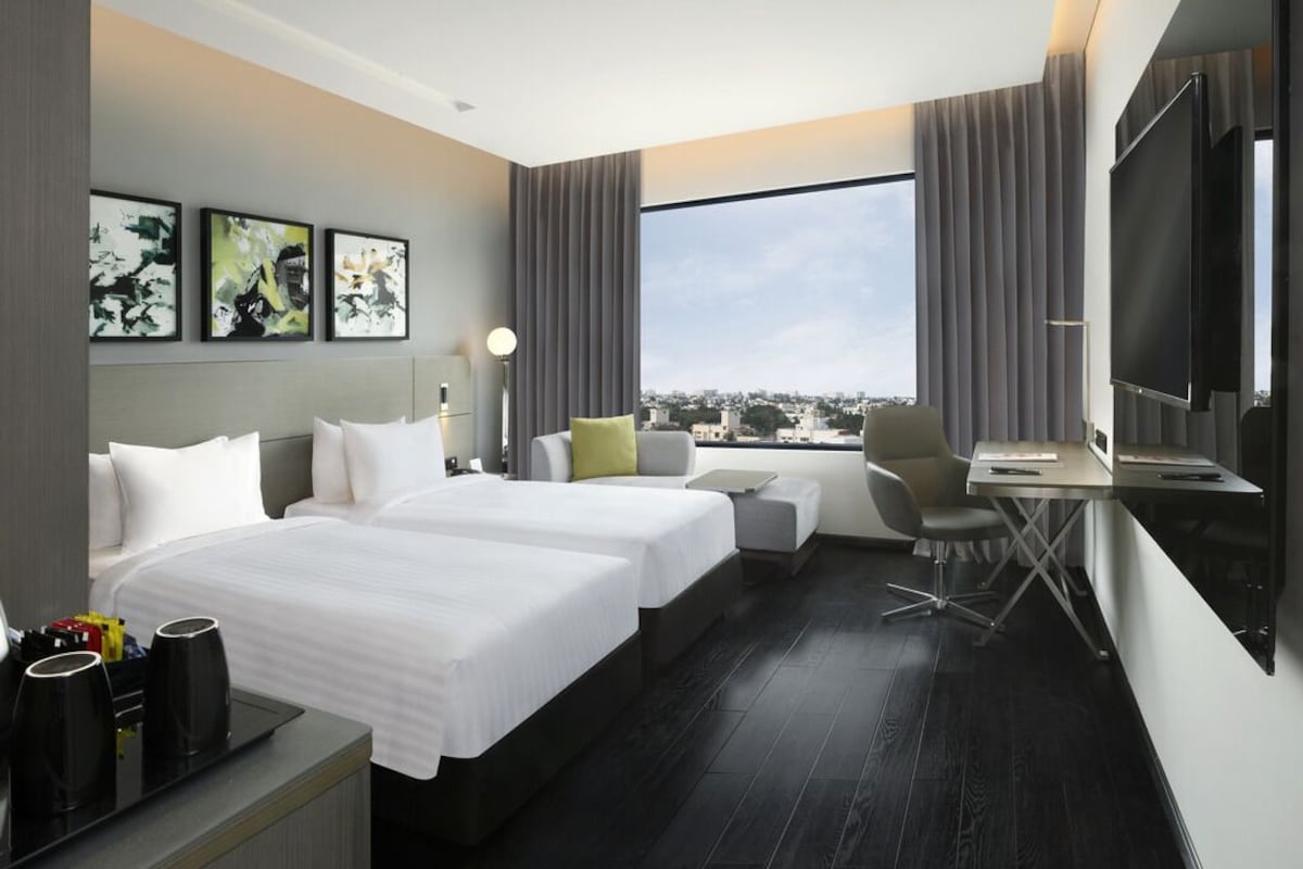 Quarto de hotel com duas camas, uma escrivaninha, uma cadeira, uma TV e vista para a cidade.