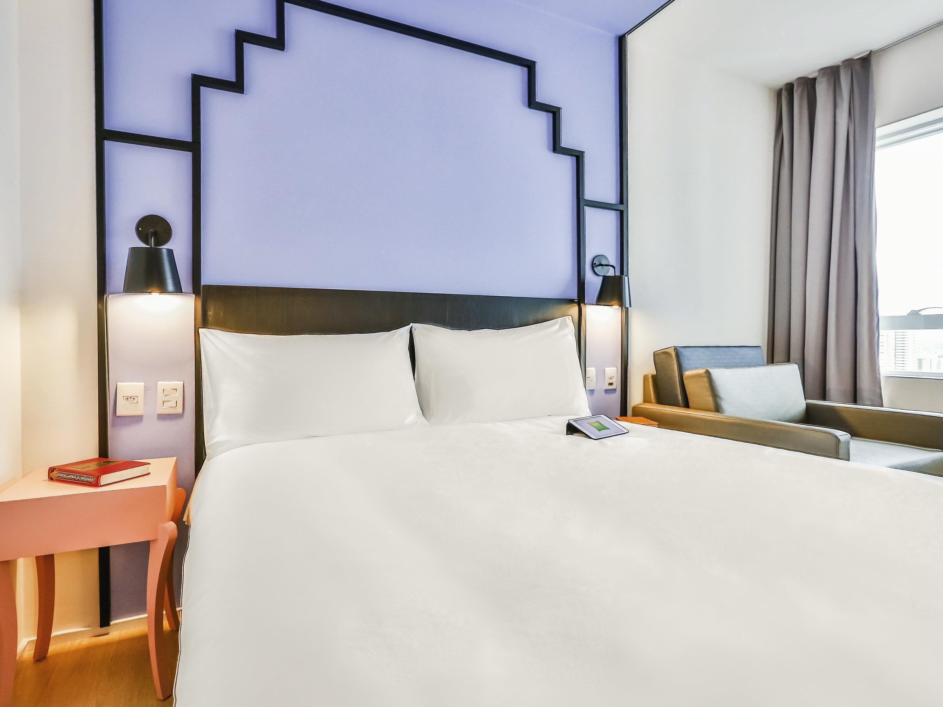 Quarto de hotel com uma cama grande, uma mesa de cabeceira com um livro, uma cadeira e uma luminária na parede.