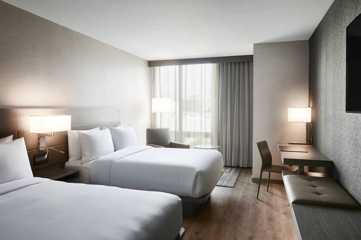 Quarto de hotel com duas camas, uma escrivaninha, uma cadeira e uma mesinha.