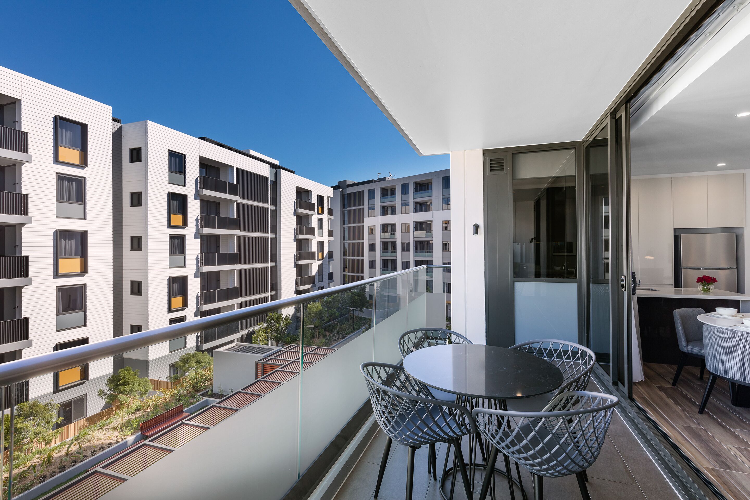 Uma varanda com guarda-corpo de vidro, mesa de jantar com cadeiras e vista para edifícios próximos.