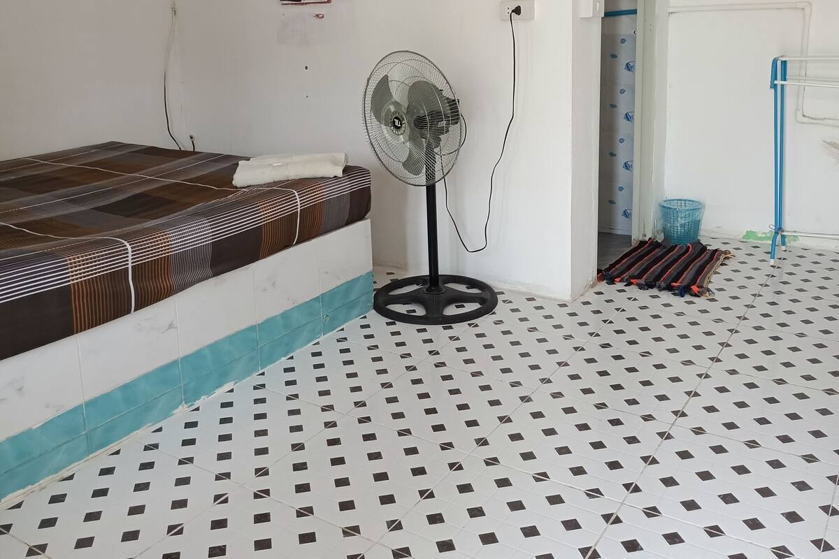 Um quarto com piso estampado, cama com colchão listrado, um ventilador e uma porta parcialmente visível que dá para outro cômodo.