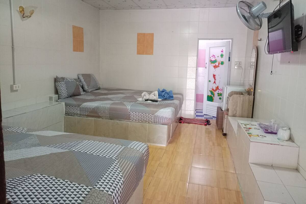 Quarto com duas camas, uma escrivaninha, uma cadeira, uma televisão e um ventilador.