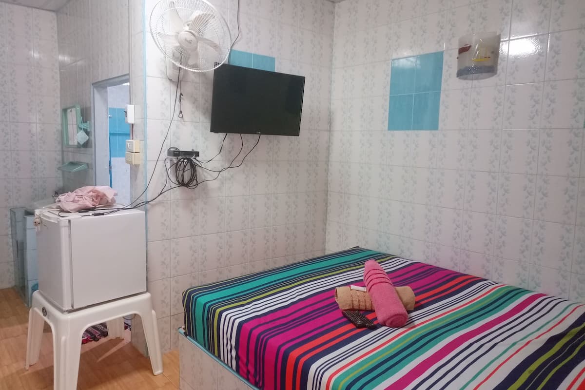 Quarto pequeno e simples com cama, uma mesinha branca, um ventilador e uma TV fixada na parede.