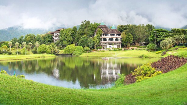 Vista serena de um lago, com uma casa grande cercada por vegetação exuberante e um lago.