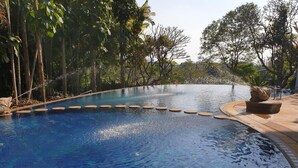 Uma piscina infinita transparente, com curvas, rodeada por vegetação tropical e um deck de pedra.
