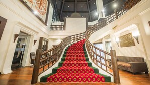 Uma escadaria imponente com tapete vermelho, guarda-corpos de madeira e teto alto com claraboias.
