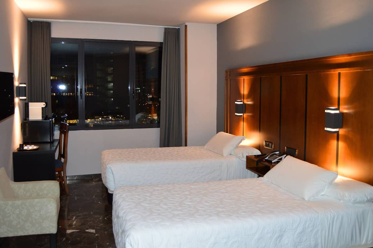 Quarto de hotel com duas camas, uma escrivaninha, uma cadeira e uma janela grande com vista para a cidade à noite.