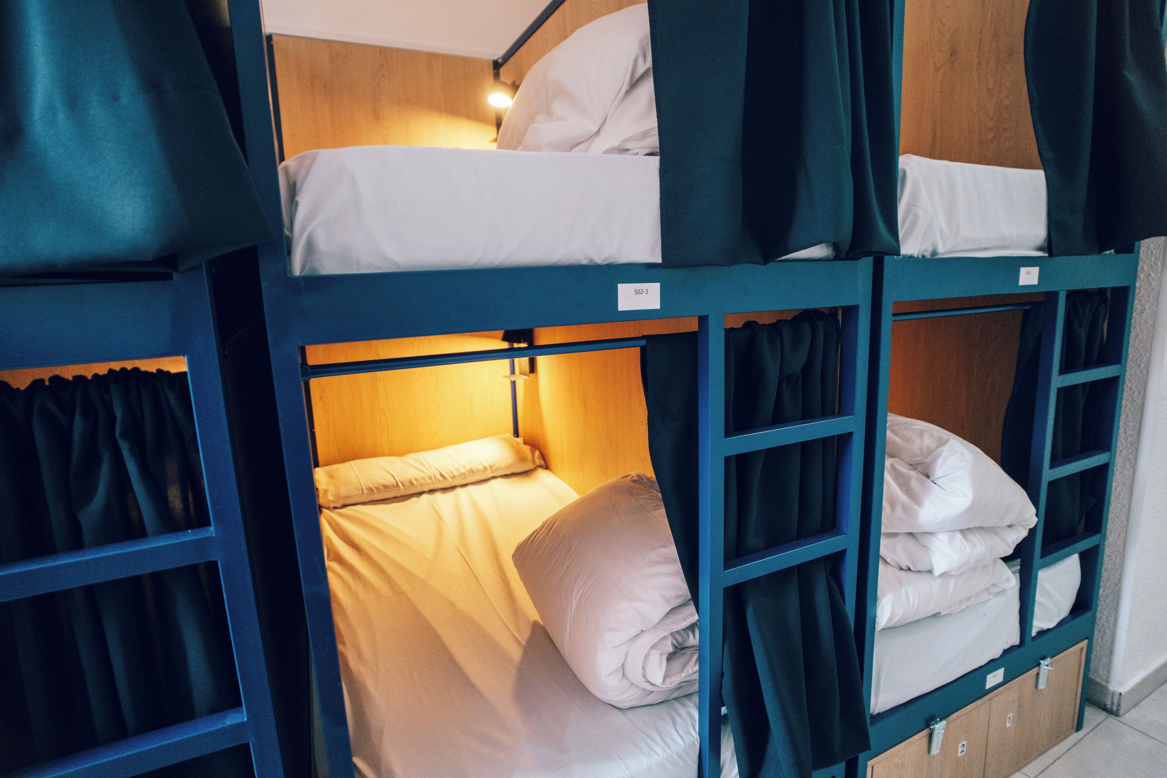 Beliches com roupa de cama branca e cortinas escuras, lambris de madeira e uma pequena prateleira com roupas dobradas.