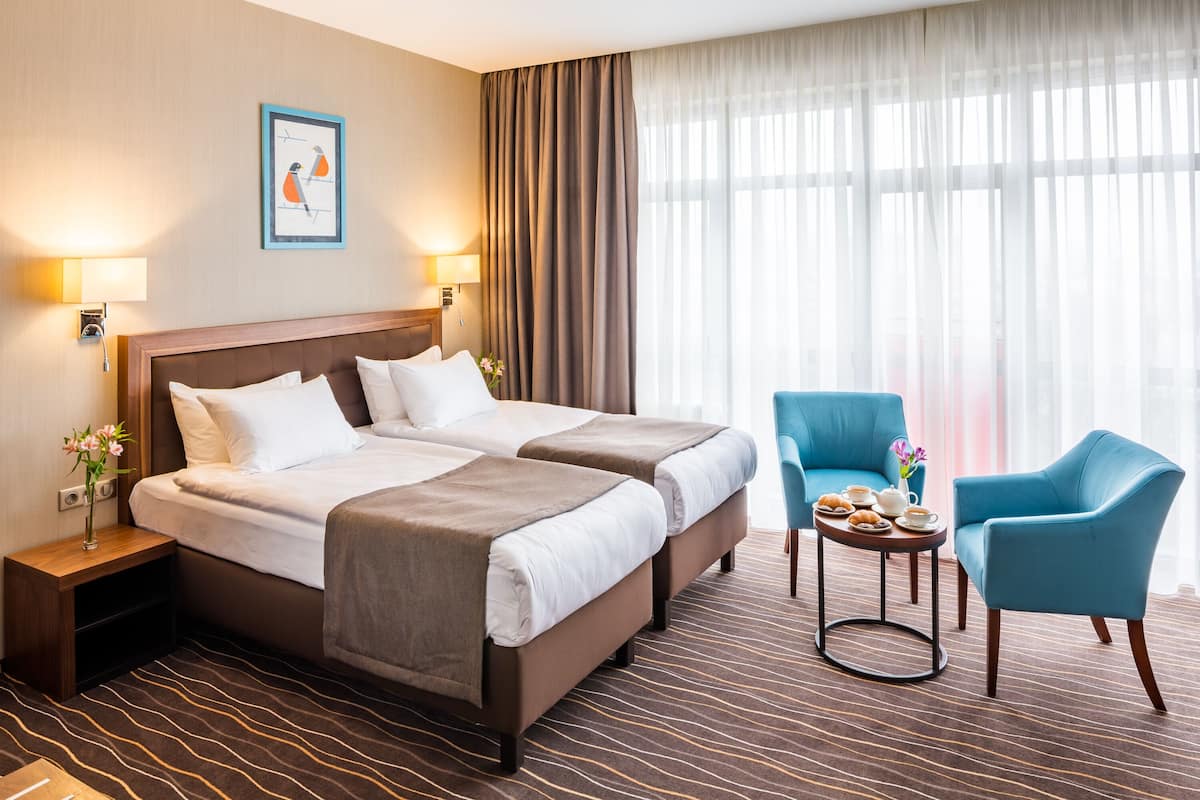Quarto de hotel com cama grande, duas mesas de cabeceira, uma mesinha redonda com bule e doces, uma poltrona azul e uma janela ampla com cortinas leves.