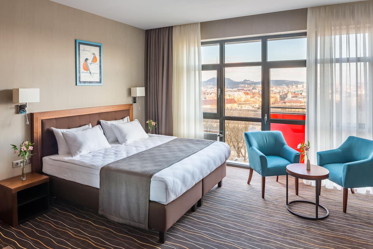Quarto de hotel com uma cama grande, duas cadeiras, uma mesinha e vista para a paisagem urbana.