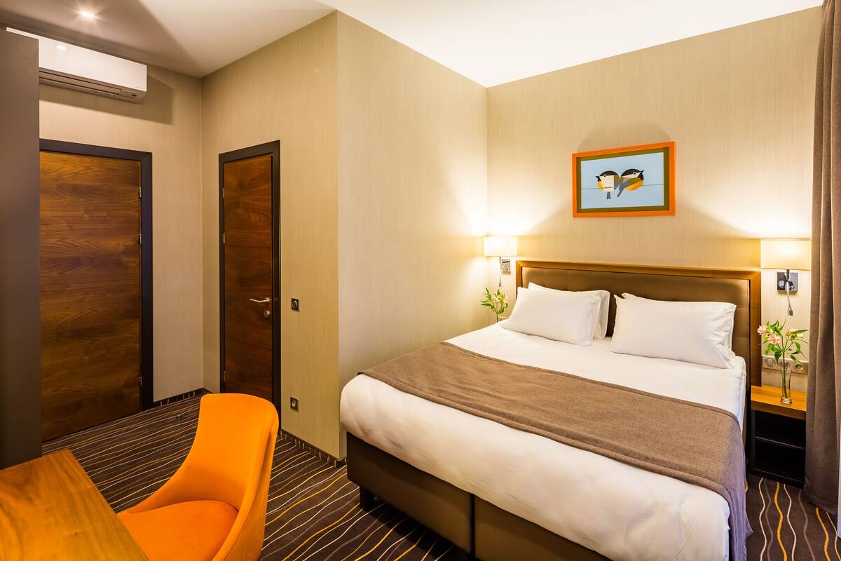Quarto de hotel com cama grande, porta de madeira, cadeira laranja e quadro na parede.
