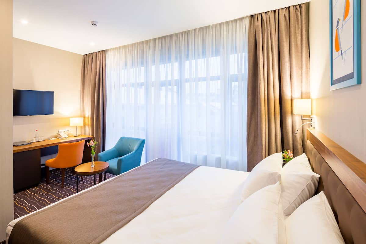 Quarto de hotel com cama, escrivaninha com cadeira, televisão e janela com cortinas.