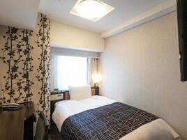 Quarto de hotel com cama, escrivaninha, telefone, mesa de cabeceira e janela com cortinas.