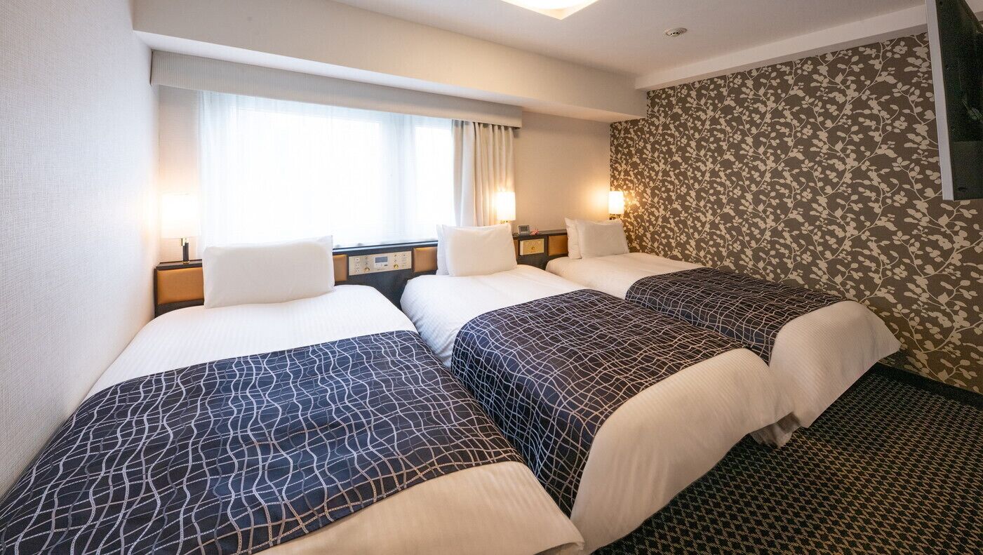 Quarto de hotel com duas camas, uma janela com cortinas, parede com padrão floral e luminárias de cabeceira.