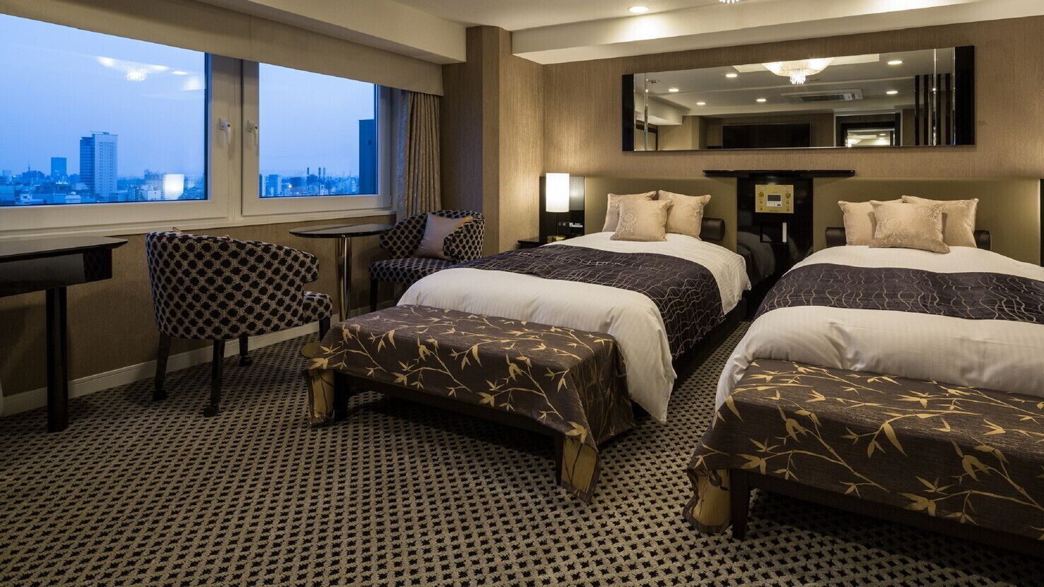 Quarto de hotel com duas camas, uma escrivaninha, uma cadeira e uma janela grande com vista para a cidade.