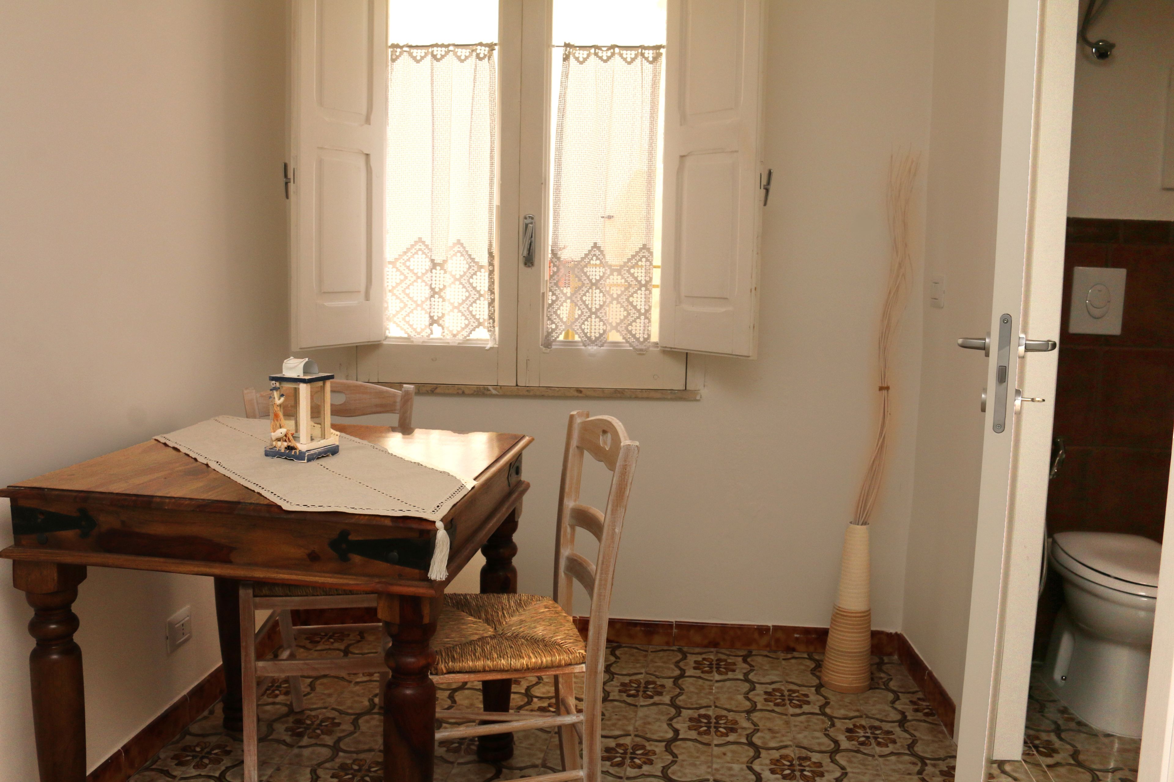 Uma pequena área de refeições com mesa e cadeiras de madeira, uma janela com cortinas de renda e uma vassoura encostada na parede.