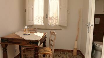Uma pequena área de refeições com mesa e cadeiras de madeira, uma janela com cortinas de renda e uma vassoura encostada na parede.