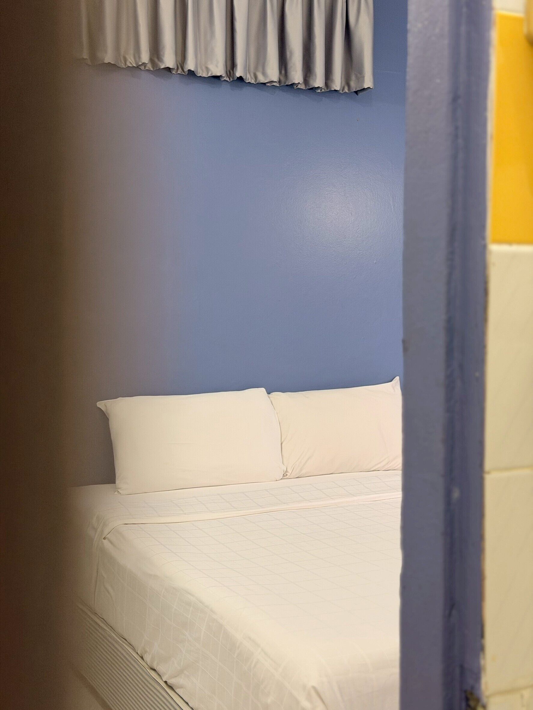 Cama com roupa de cama branca, parede azul e parte de um batente de porta visível.