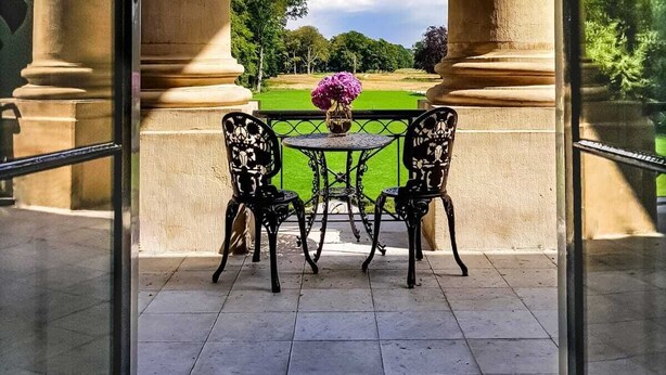 Varanda com mesa e cadeiras, um vaso de flores e vista para uma paisagem verde através de grandes janelas emolduradas por colunas.
