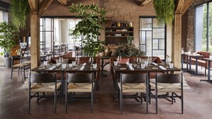 Restaurante com vigas de madeira, janelas amplas e plantas penduradas. Mesas montadas com copos de vinho e pratos.