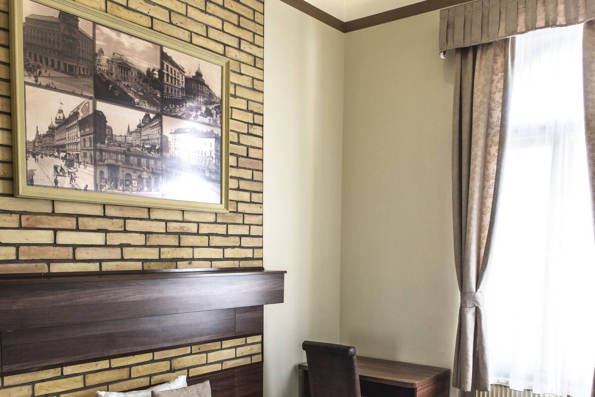 Uma cama bem arrumada com cabeceira, uma mesa de trabalho e uma cadeira num quarto com parede de tijolos e uma janela grande com cortinas.
