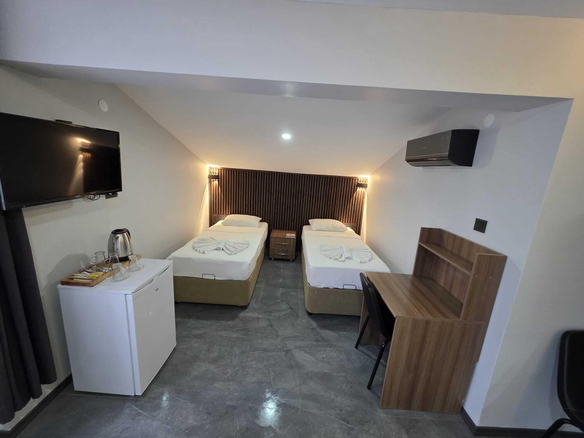 Quarto de hotel compacto com duas camas de solteiro, uma kitchenette pequena, uma mesa e uma televisão.