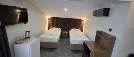 Quarto de hotel compacto com duas camas de solteiro, uma kitchenette pequena, uma mesa e uma televisão.