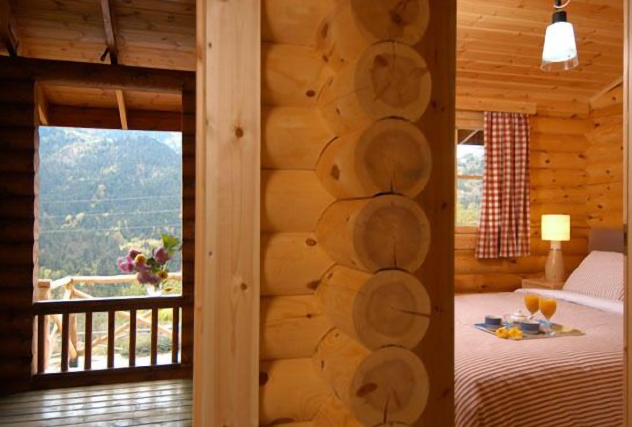 Quarto de cabana de madeira com cama, mesa de cabeceira, abajur e janela com vista para montanhas.