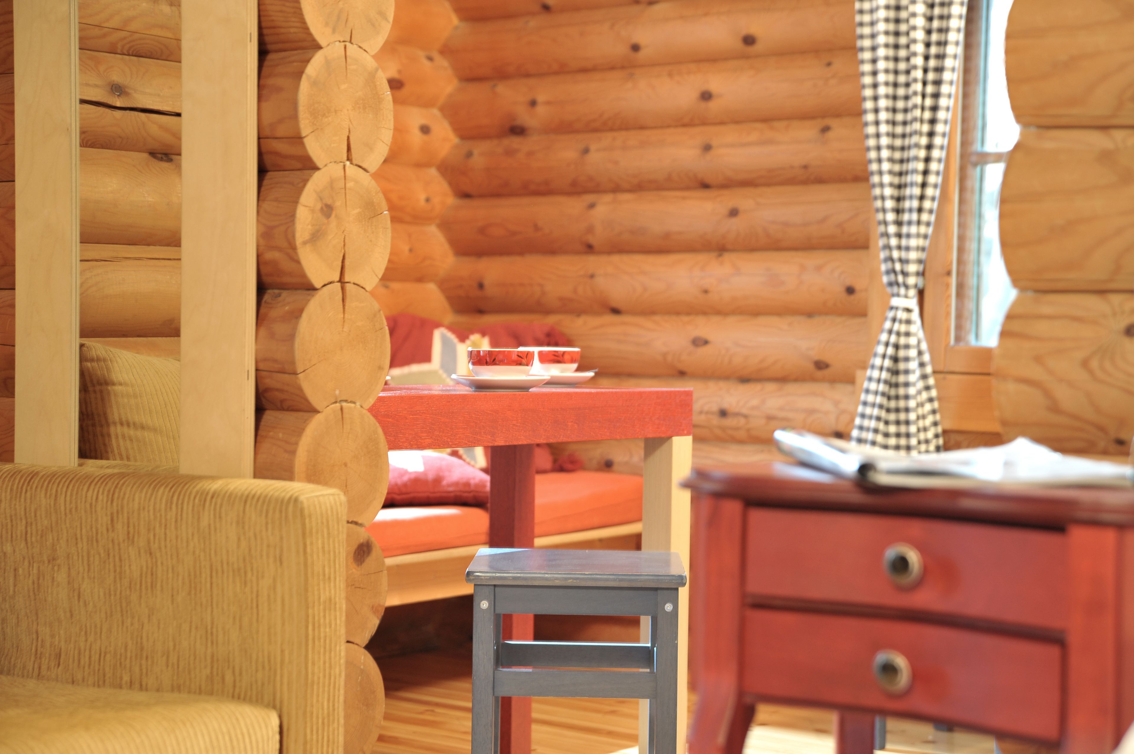 Interior de cabana de tronco com parede de madeira, mesa vermelha com prato branco, cortina xadrez e um banquinho pequeno.