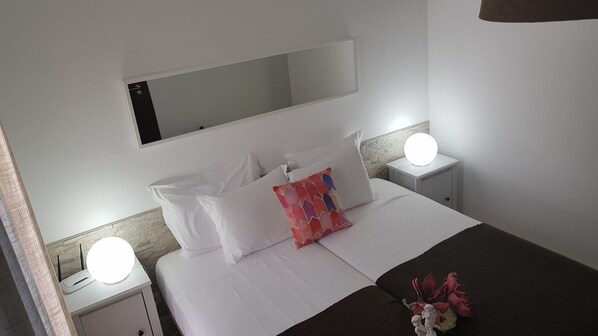 Quarto de hotel com cama bem arrumada, roupa de cama branca, almofada decorativa, mesa de cabeceira com abajur e espelho fixado na parede.