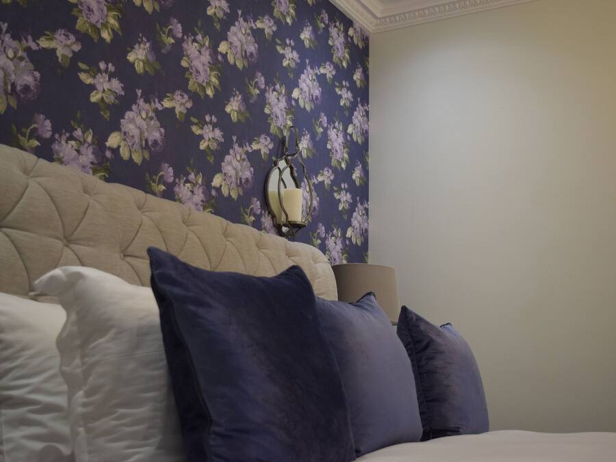 Quarto com cabeceira estofada, cama com almofadas brancas e azul-escuras, parede com papel de parede floral e abajur.