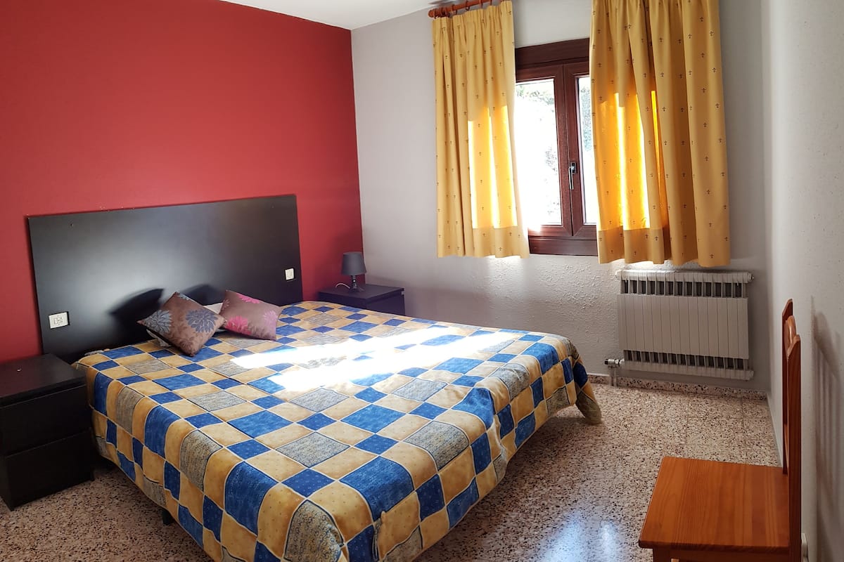 Quarto com colcha xadrez, mesa de cabeceira com abajur, radiador e janela com cortinas.