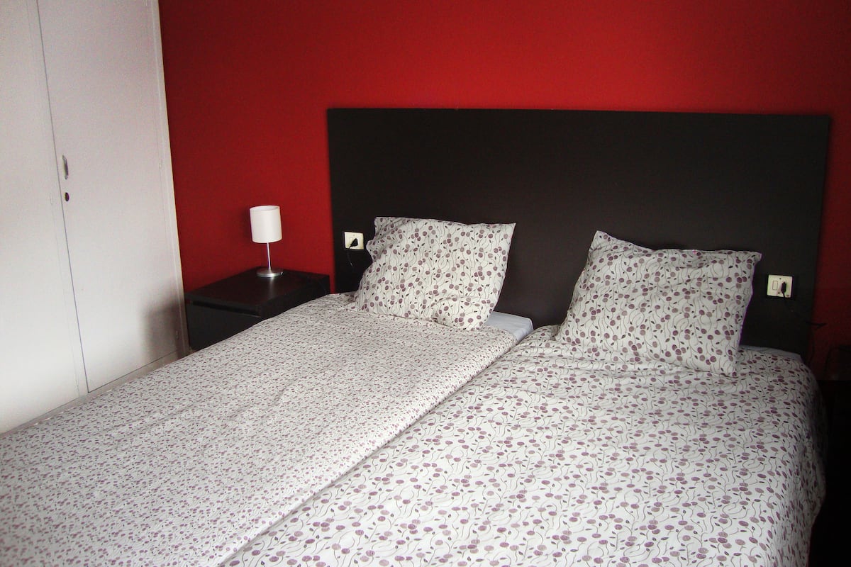 Cama com roupa de cama florida, mesa de cabeceira com abajur e parede vermelha.