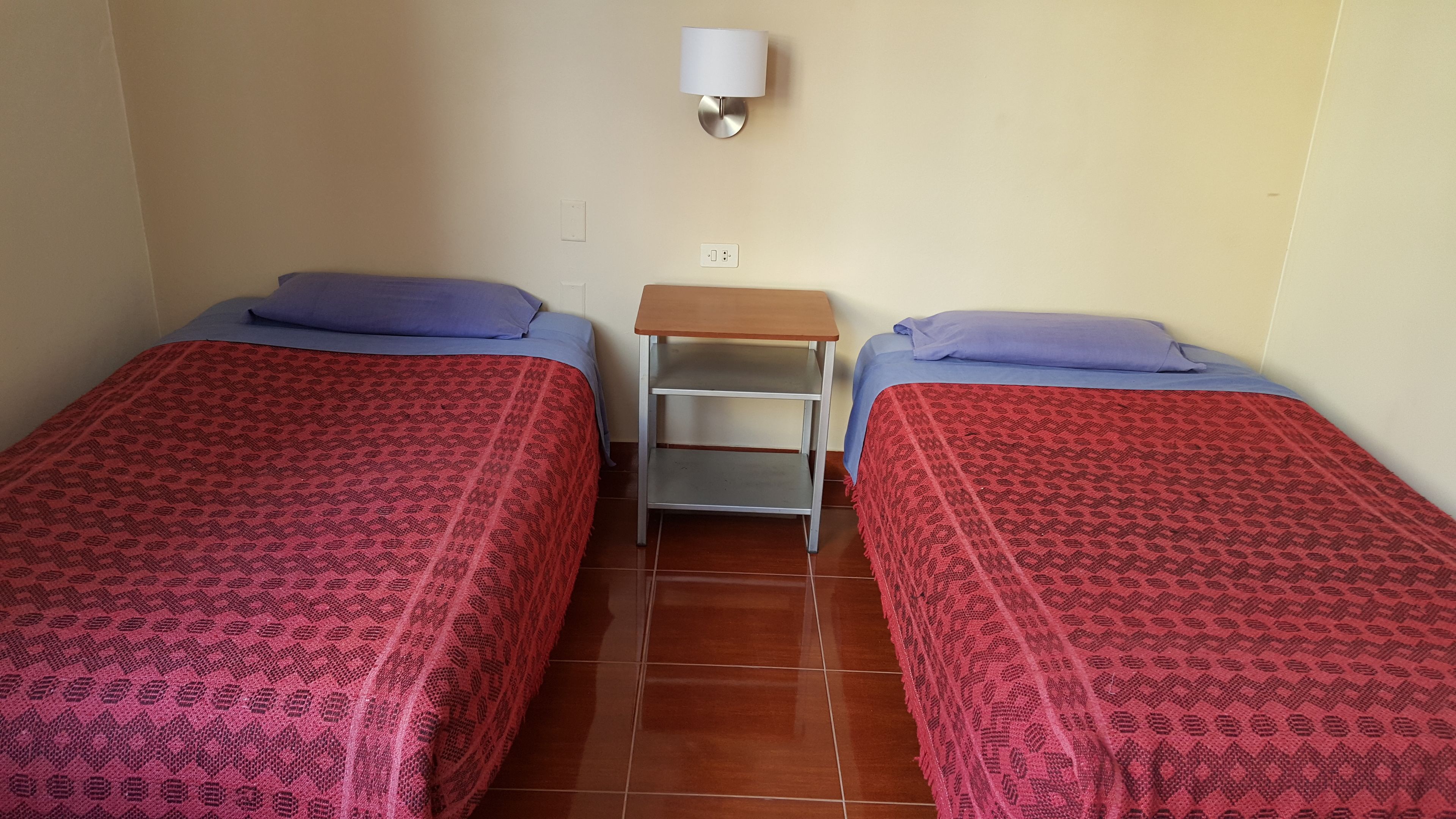 Quarto com duas camas, cada uma com colcha vermelha estampada e travesseiro azul. Há uma pequena mesa de madeira entre as camas e um abajur fixado na parede acima.
