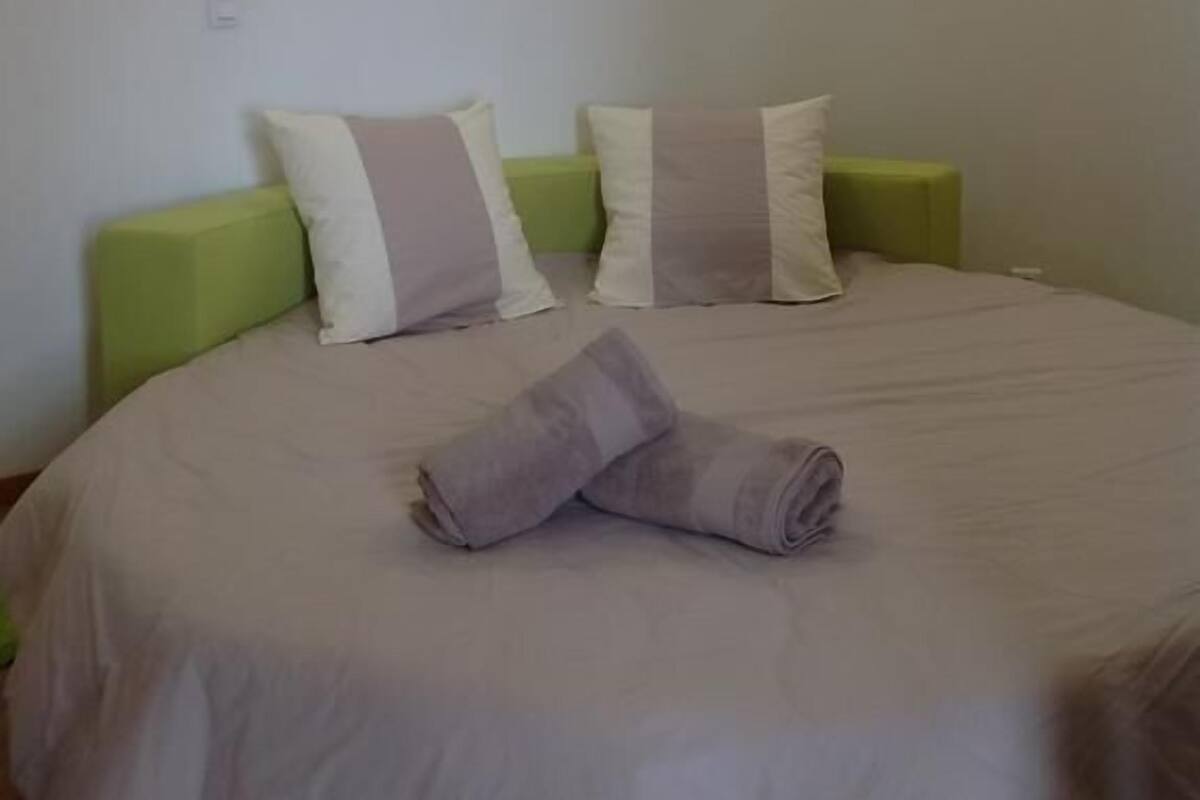 Cama redonda com colcha bege, duas almofadas e uma toalha dobrada. Duas luzes fixadas na parede acima da cama.