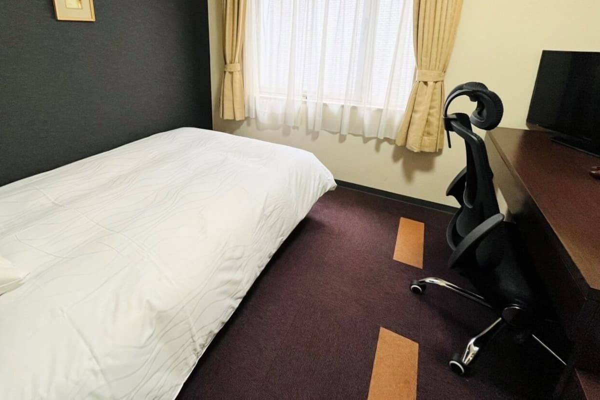Quarto de hotel com uma cama de solteiro, uma escrivaninha com cadeira, televisão e janela com cortinas.