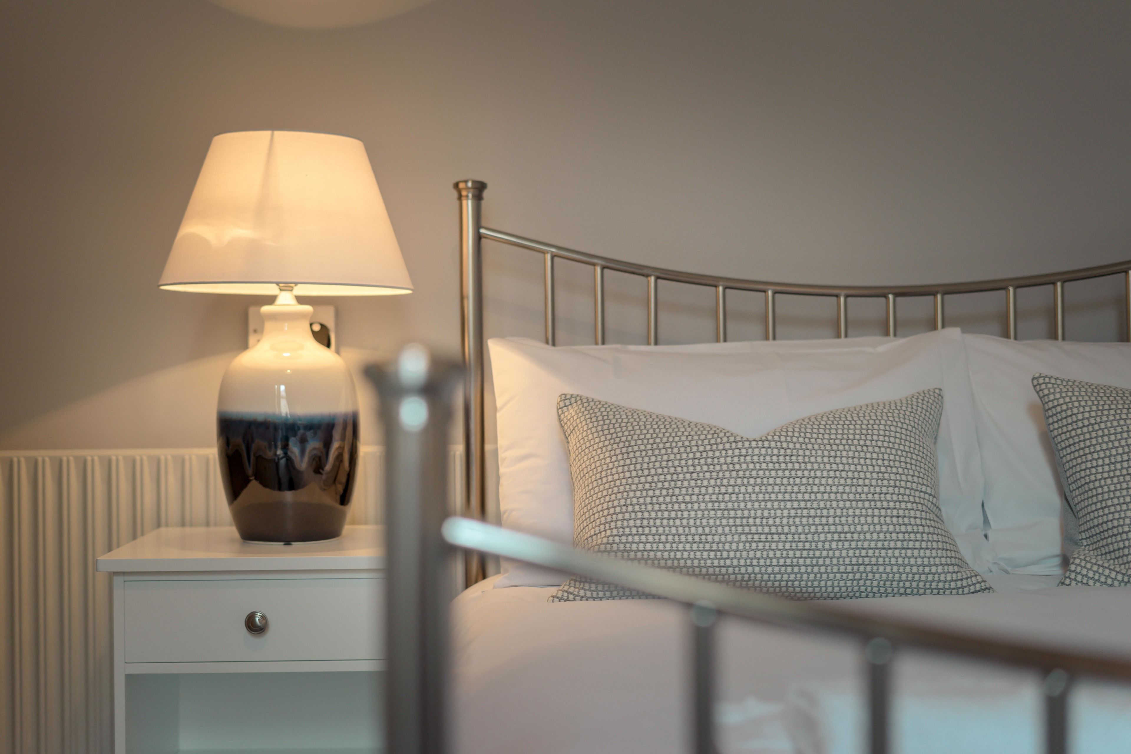Cama bem arrumada com almofadas brancas e estampadas, mesa de cabeceira branca com abajur e cabeceira de metal.