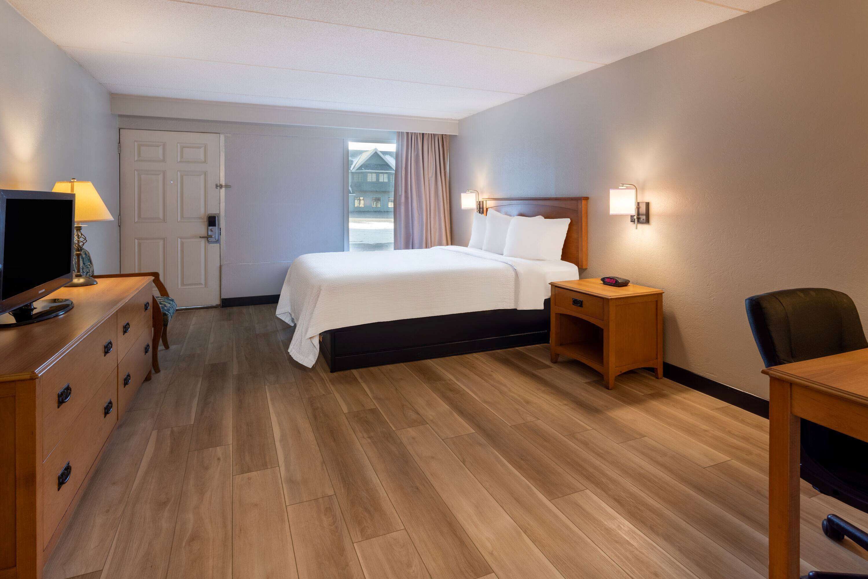 Quarto de hotel com cama, escrivaninha, cadeira, televisão e porta.